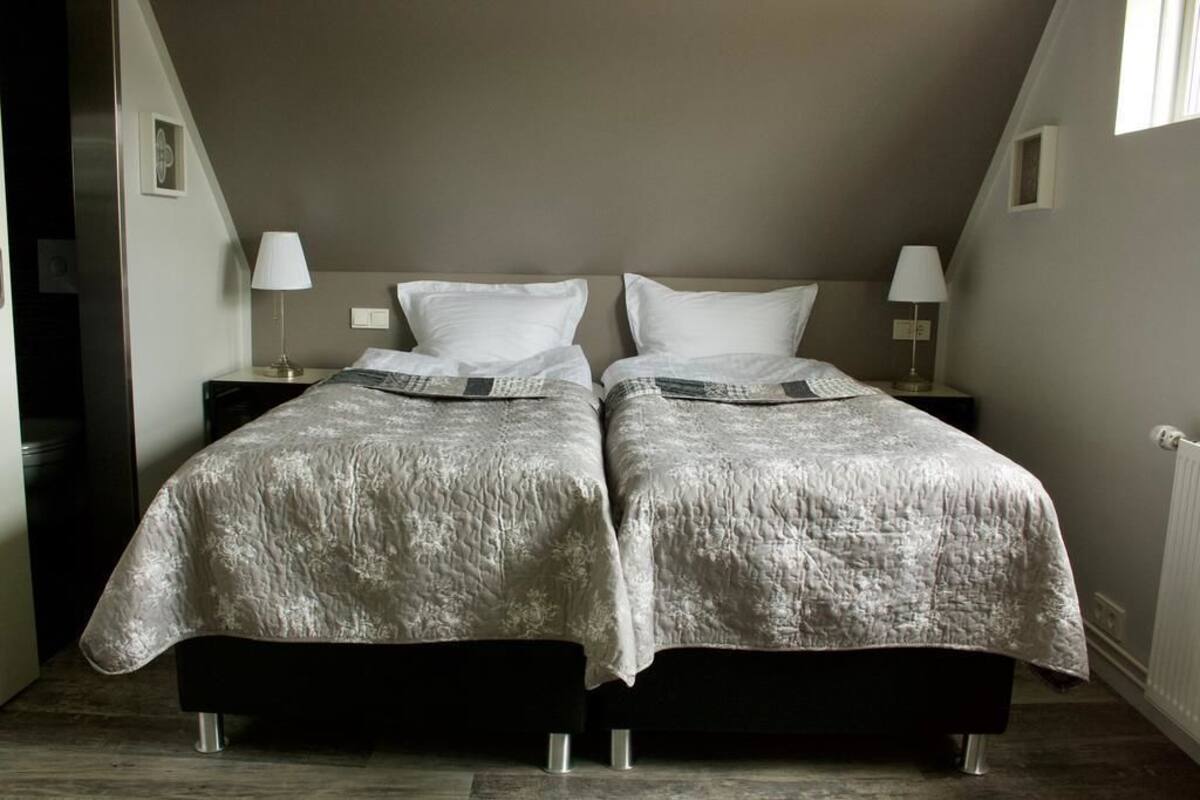 Quarto duplo com duas camas, cada uma com travesseiro branco e cabeceira combinando. Há mesas de cabeceira com abajures e um radiador na parede.