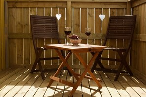 Duas cadeiras de madeira e uma mesa com uma tigela de uvas e duas taças de vinho tinto, dispostas diante de um cercado de madeira com desenhos em formato de coração.