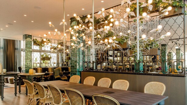 Restaurante com uma longa mesa de madeira e cadeiras de vime, área de bar com azulejos verdes e luminárias pendentes com folhas.