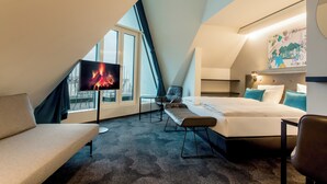 Quarto de hotel moderno com uma cama grande, um sofá, uma TV e vista da cidade através de uma janela ampla.