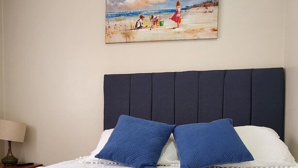 Cama bem arrumada com roupa de cama azul e branca, um quadro de uma paisagem de praia na parede e um abajur ao lado.