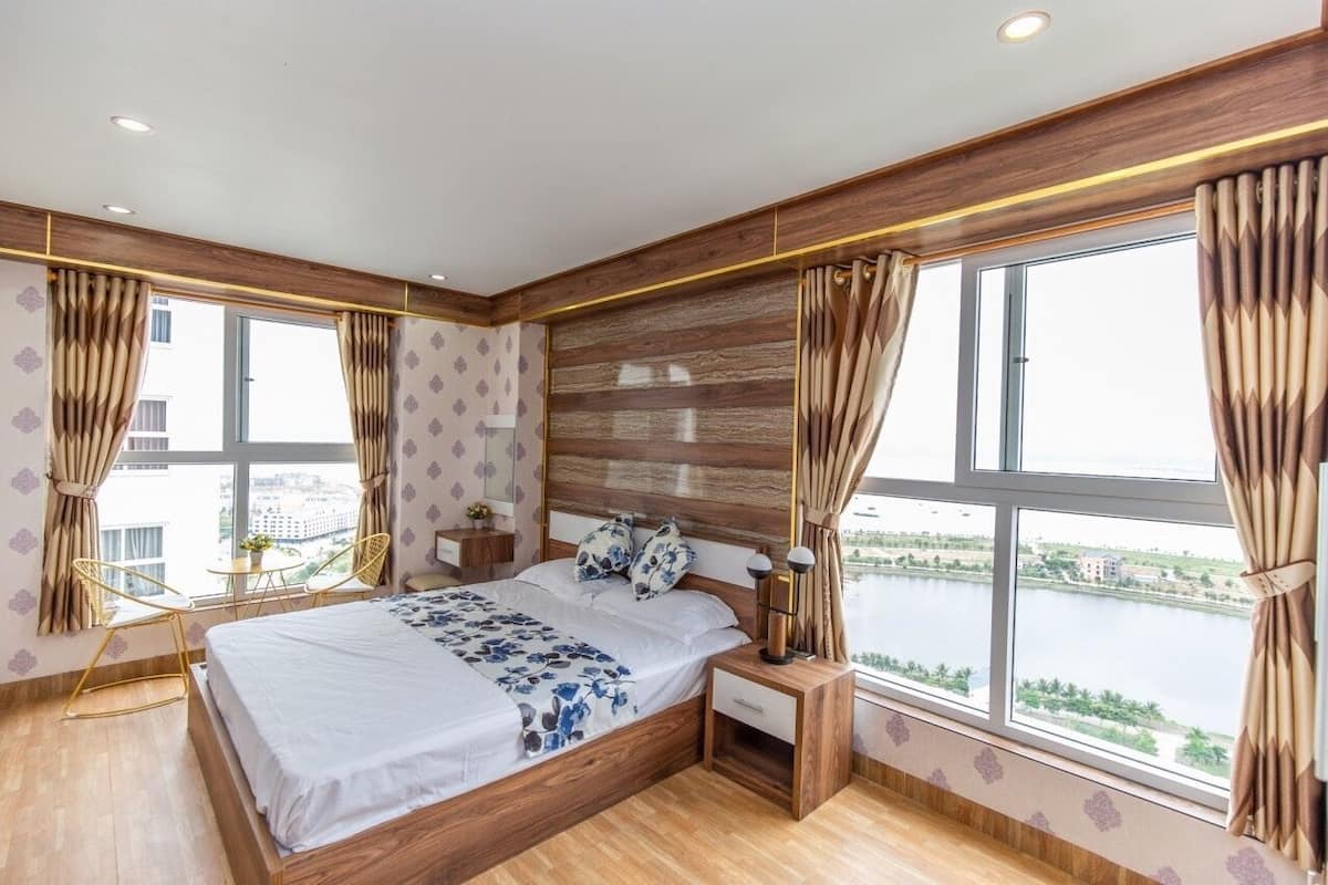 Quarto com cama grande, duas mesas de cabeceira, janela com cortinas e vista para a água.