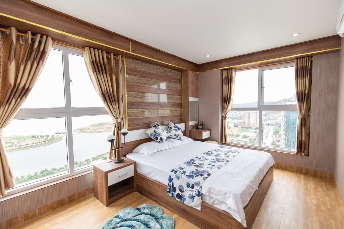 Quarto com uma cama grande, duas mesas de cabeceira, um criado-mudo, uma janela com cortinas e vista para um corpo d'água.