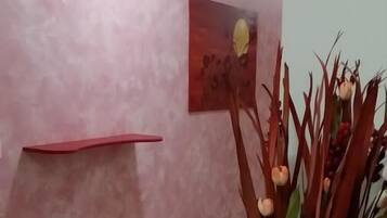 Um corredor com parede rosa, piso ladrilhado, prateleira vermelha e um vaso com plantas decorativas.