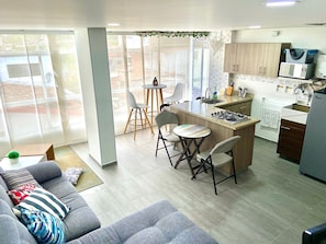 Cozinha moderna com ilha central, sala de jantar com cadeiras e sala de estar com sofá e almofadas.