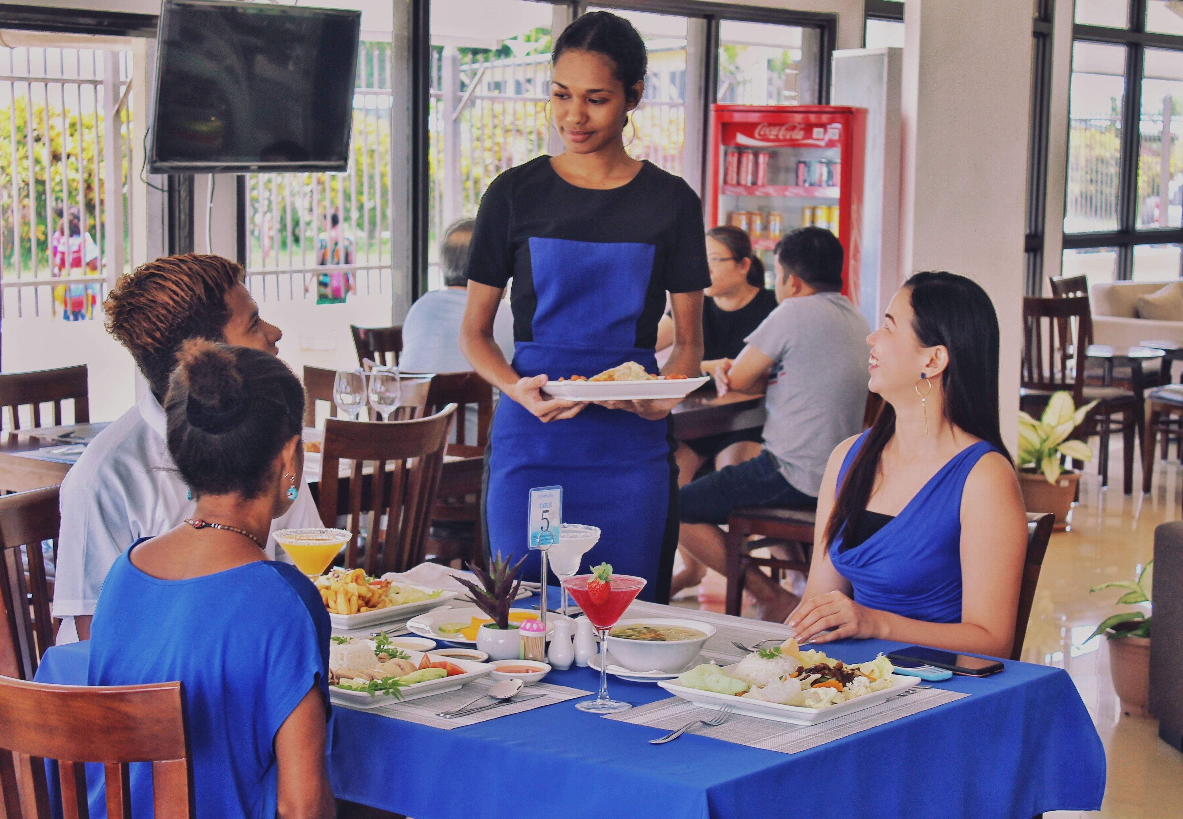 Um restaurante com toalha de mesa azul, um refrigerador de Coca-Cola e uma televisão na parede.