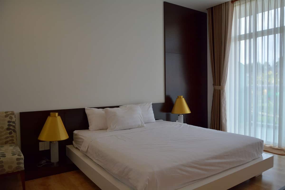 Quarto de hotel moderno com uma cama grande, duas luminárias de cabeceira, uma mesinha de noite e vista para o exterior através de cortinas translúcidas.