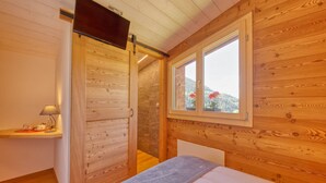 Quarto de cabana de madeira com cama, janela com vista para flores vermelhas e mesa com abajur e xícaras.