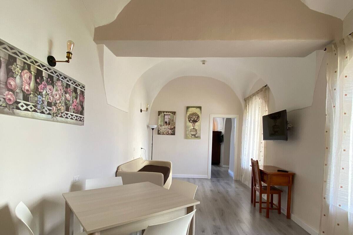 Uma área de refeições com uma mesa e cadeiras de madeira, uma TV fixada na parede e um corredor que dá para outro quarto.
