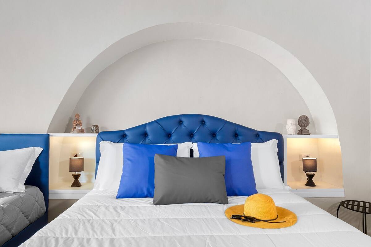 Uma cama com cabeceira azul, travesseiros brancos e azuis, um travesseiro cinza, uma colcha branca, um chapéu amarelo e duas luminárias de mesa.