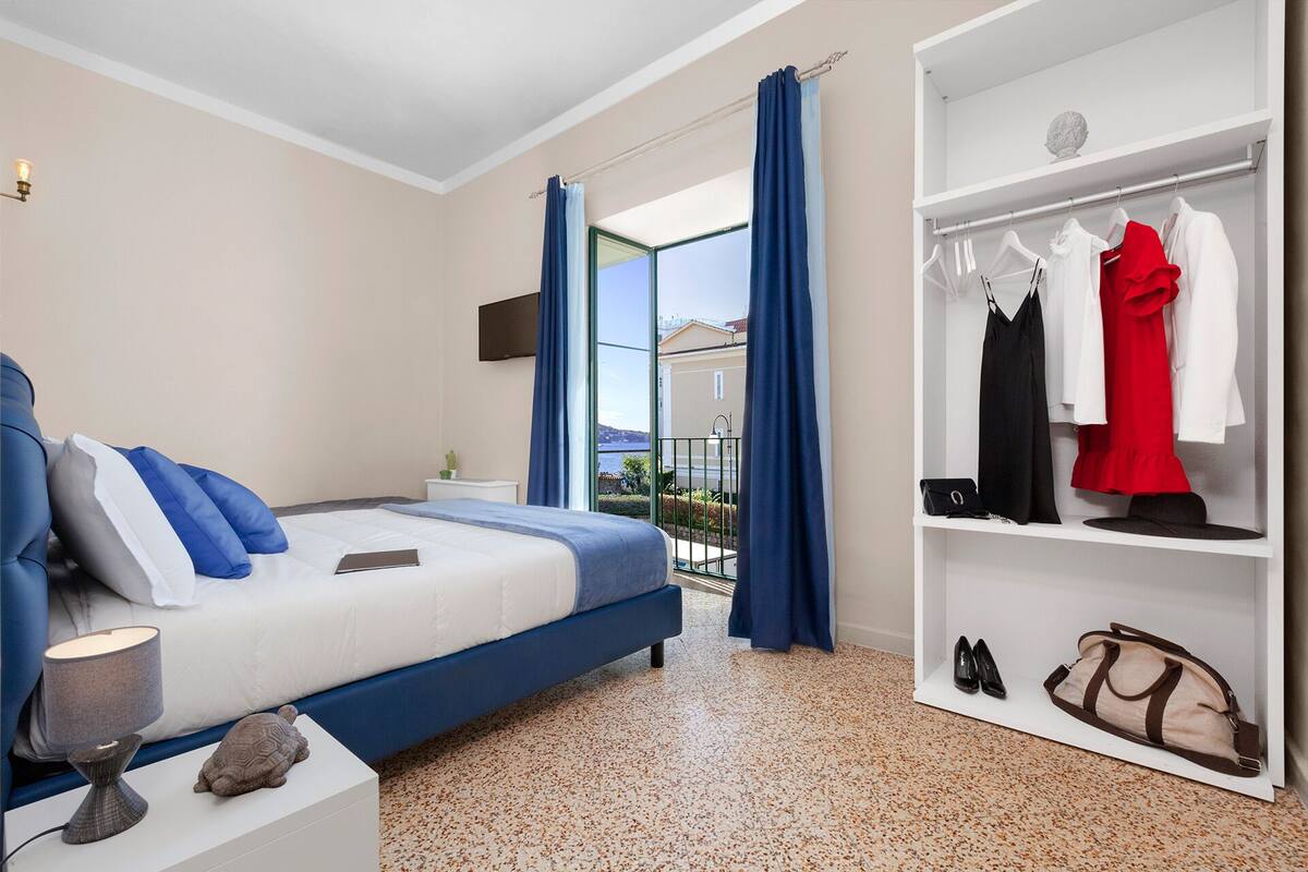 Um quarto com uma cama, uma mesinha de cabeceira com abajur, uma plantinha, uma janela com cortinas e vista para o exterior.