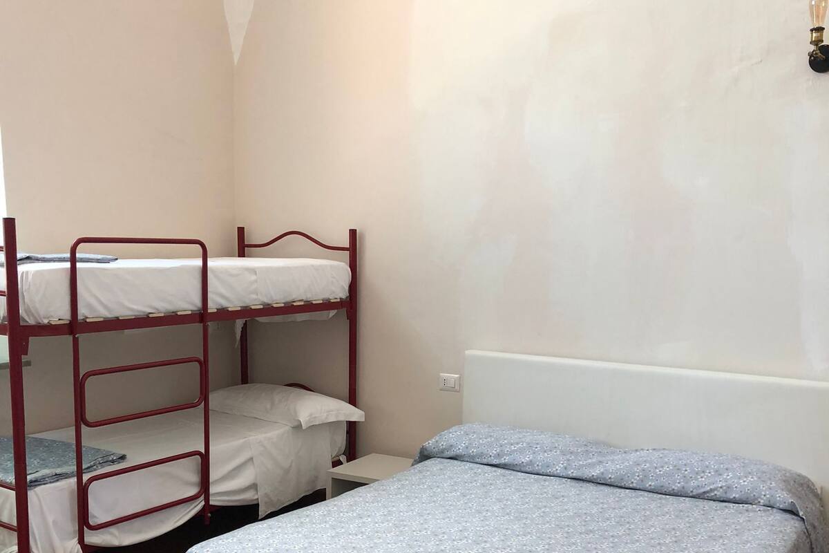Quarto tipo dormitório com beliches, uma cama de solteiro e uma parede branca com interruptor de luz.