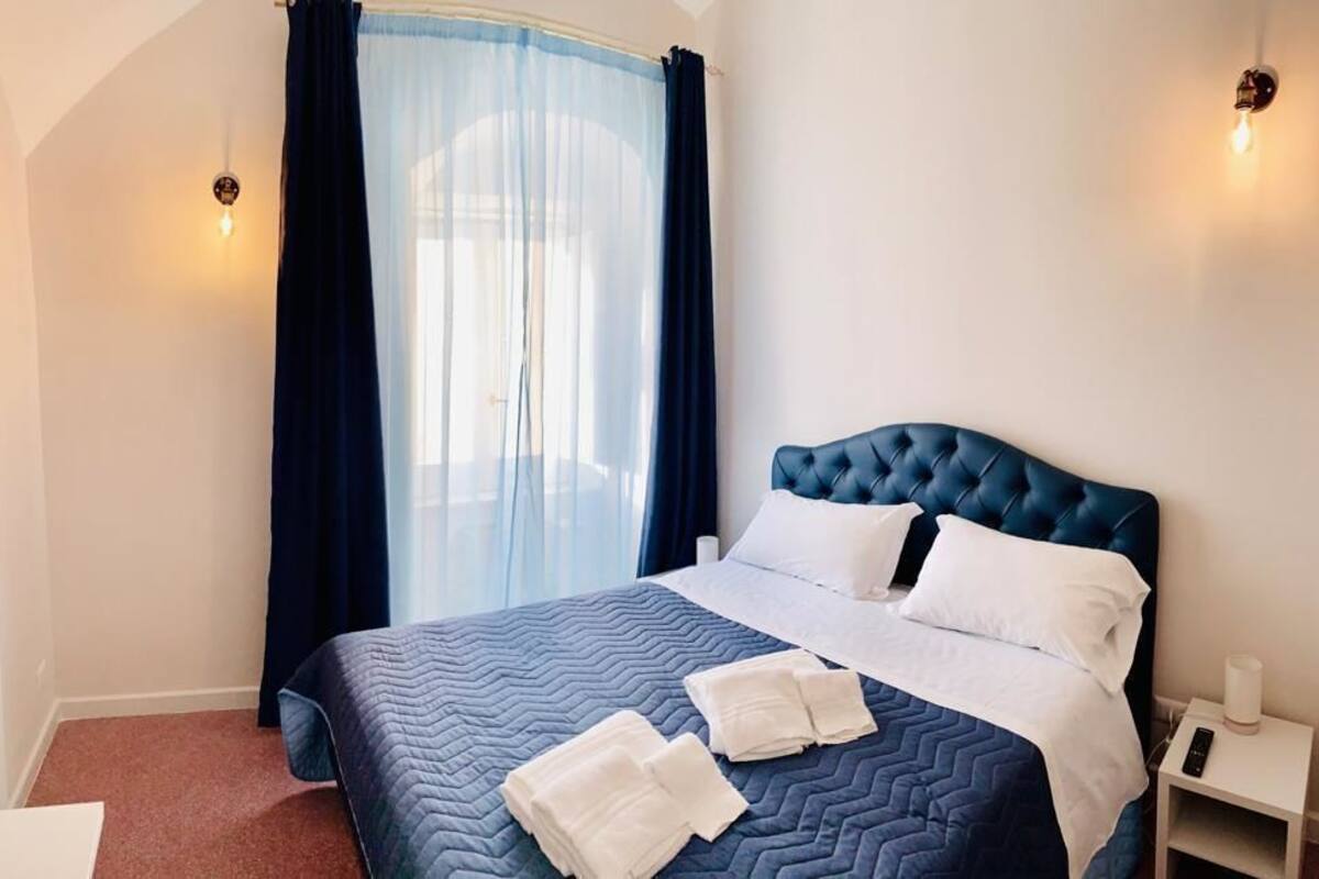 Quarto de hotel com cama, mesa de cabeceira, abajur e janela com cortinas.
