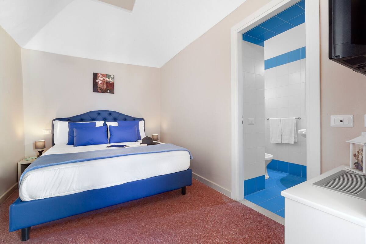 Quarto com cama, mesas de cabeceira, um quadro e banheiro com chuveiro e vaso sanitário.