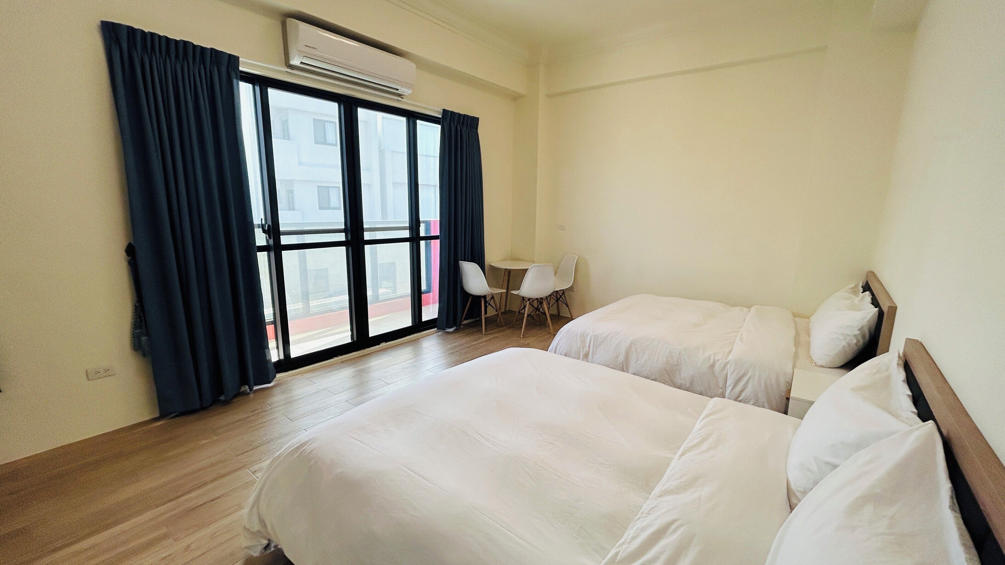 Quarto de hotel com duas camas, uma mesinha com cadeiras e uma janela grande com cortinas.