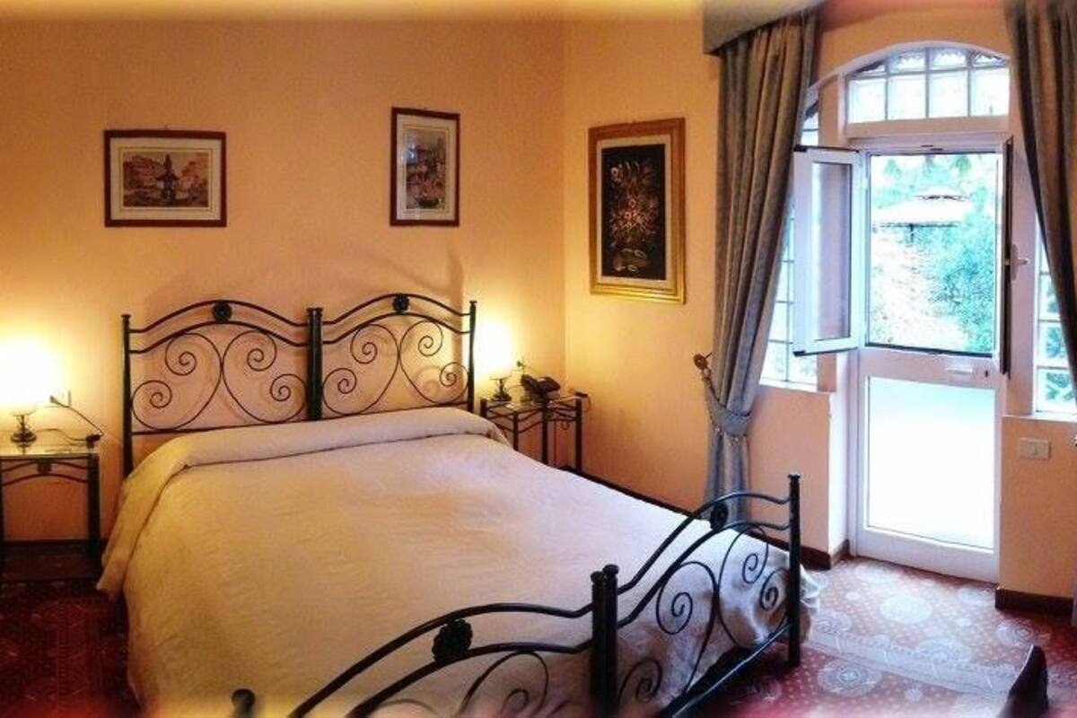Quarto com cama, mesa de cabeceira, espelho, janela com cortinas e vaso com flores.