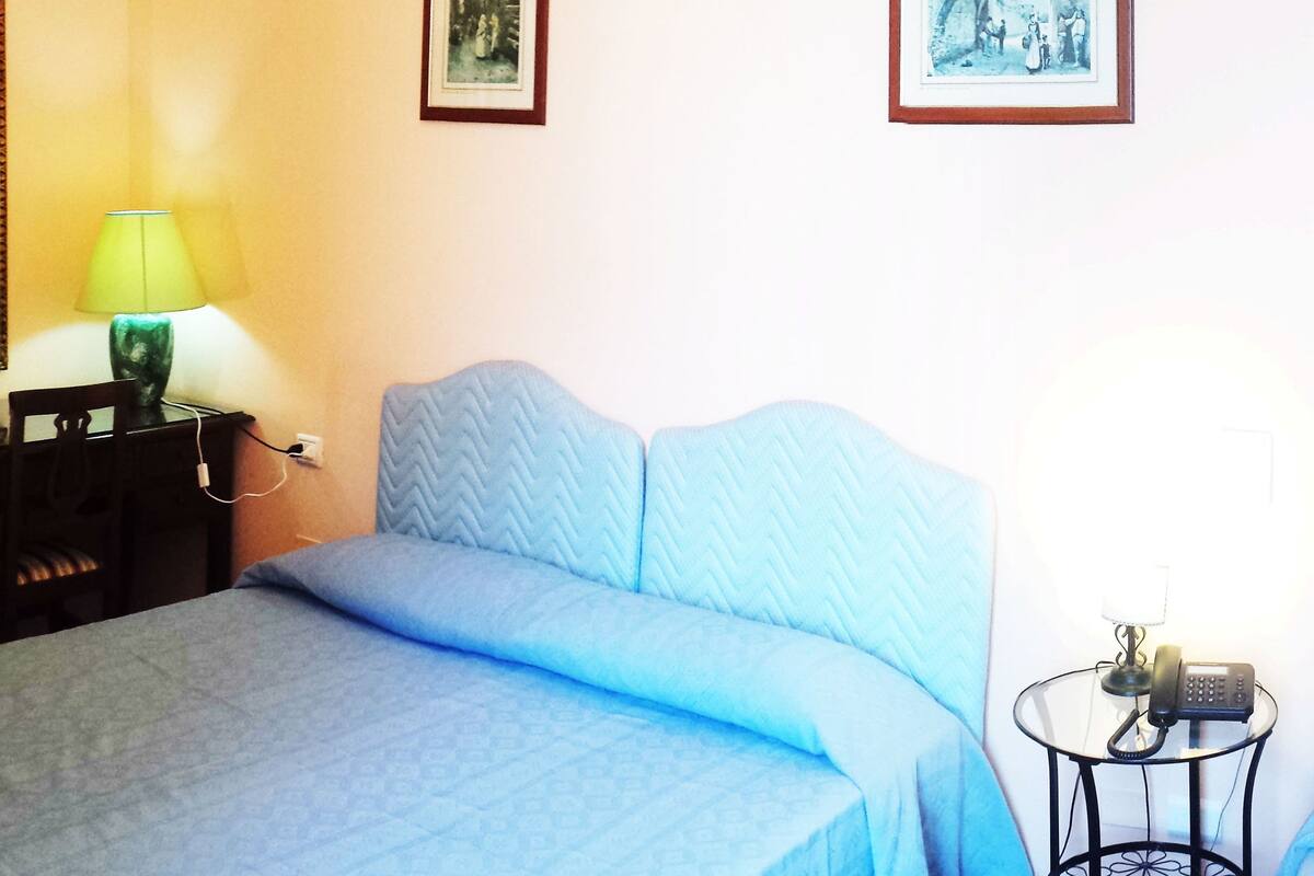 Quarto de hotel com uma cama, duas poltronas azuis estofadas, uma mesa pequena com um telefone, um abajur e um espelho.