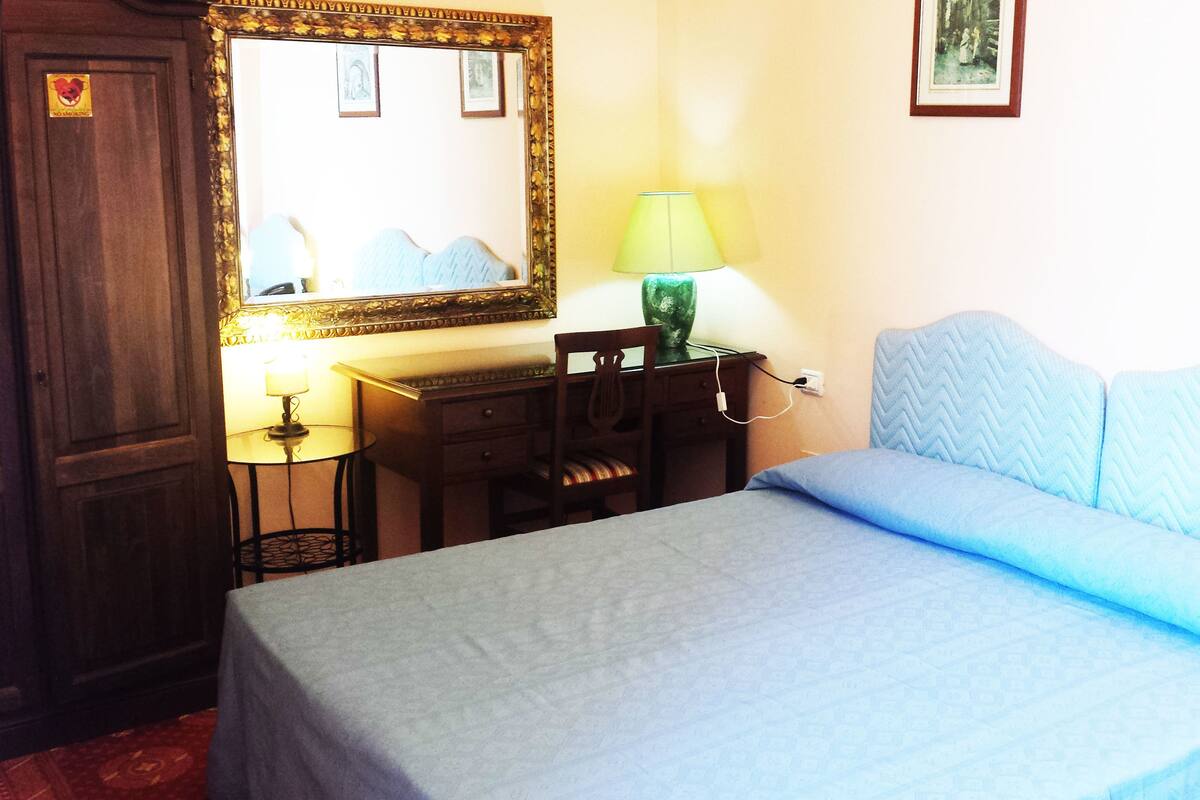 Quarto com cama, escrivaninha, cadeira, abajur e espelho.