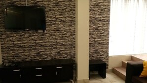 Uma sala de estar com parede estampada em pedra, uma TV de tela plana montada sobre um móvel de entretenimento escuro e um sofá com almofada amarela.
