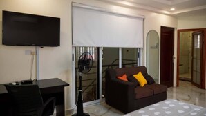Quarto de hotel com cama, escrivaninha, cadeira, televisão, janela com persiana e porta para uma casa de banho.