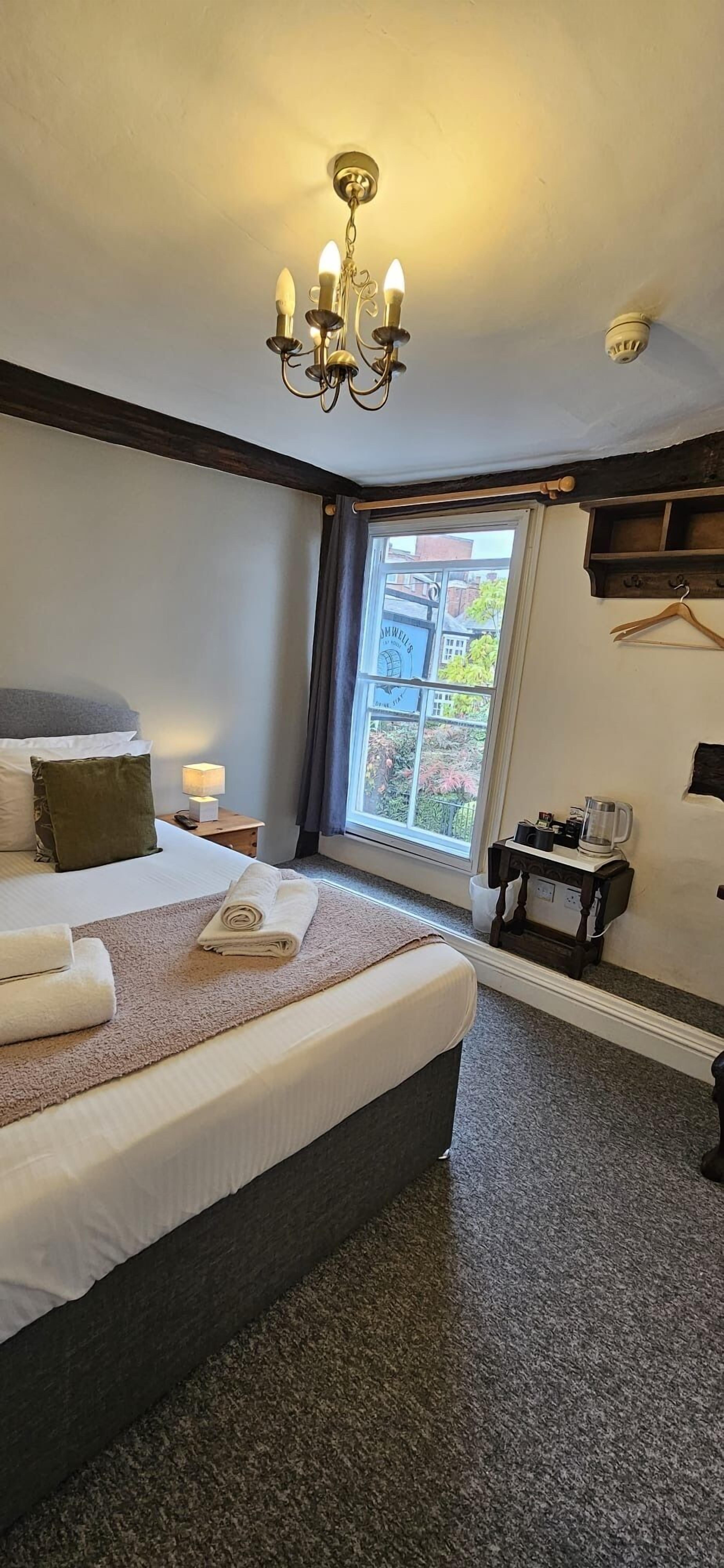 Um quarto com uma cama grande, um candelabro, uma janela com vista para prédios, uma pequena mesa com uma cafeteira e um cabideiro suspenso.