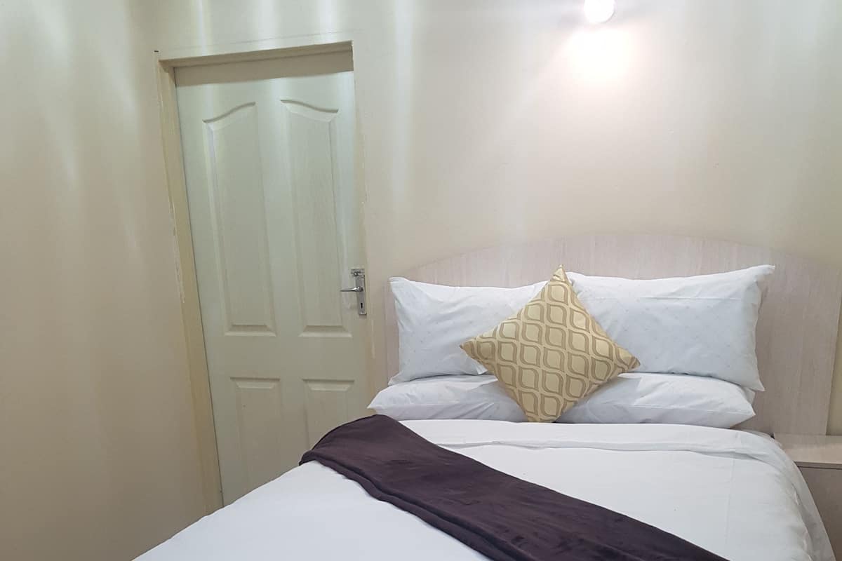 Uma cama bem arrumada com roupa de cama branca e um cobertor marrom, um tapete estampado e uma porta branca ao fundo.
