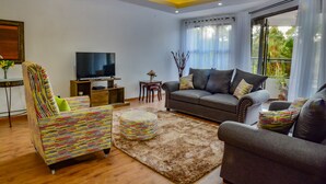 Uma sala de estar moderna com sofá, poltrona, banqueta, televisão e uma mesinha com uma planta.