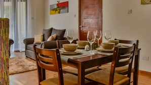 Uma sala de jantar com mesa de madeira posta para quatro pessoas, rodeada por cadeiras. Uma porta de madeira e um quadro são visíveis ao fundo.