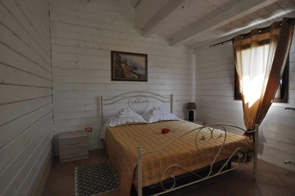 Quarto com uma cama de solteiro, mesa de cabeceira, pequeno armário e um quadro na parede, além de janela com cortinas.
