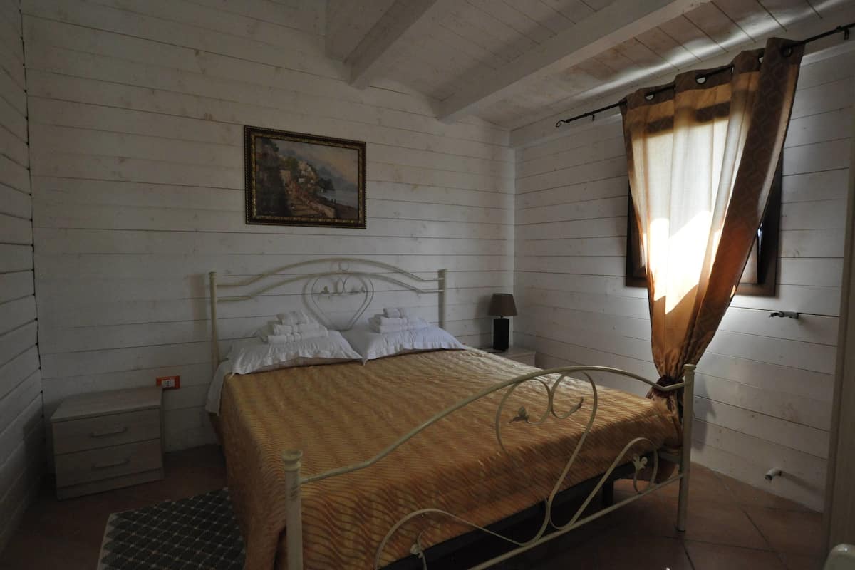 Quarto com estrutura de cama de madeira, mesa de cabeceira com abajur e um quadro na parede.