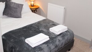 Cama bem arrumada com roupa de cama branca e cinza, luminária de cabeceira e radiador.