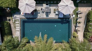 Vista aérea de uma piscina com dois grandes guarda-sóis, cercada por vegetação e um caminho pavimentado.