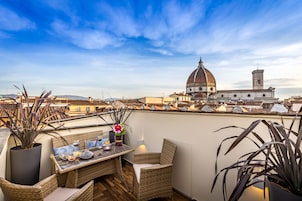 Terraço no telhado com móveis de vime, uma mesa posta para refeições e plantas em vasos. Ao fundo, visível um edifício em formato de cúpula e uma torre.