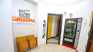 Interior de uma pensão com um letreiro com tema de cinema, uma máquina de venda automática e um banco de madeira.