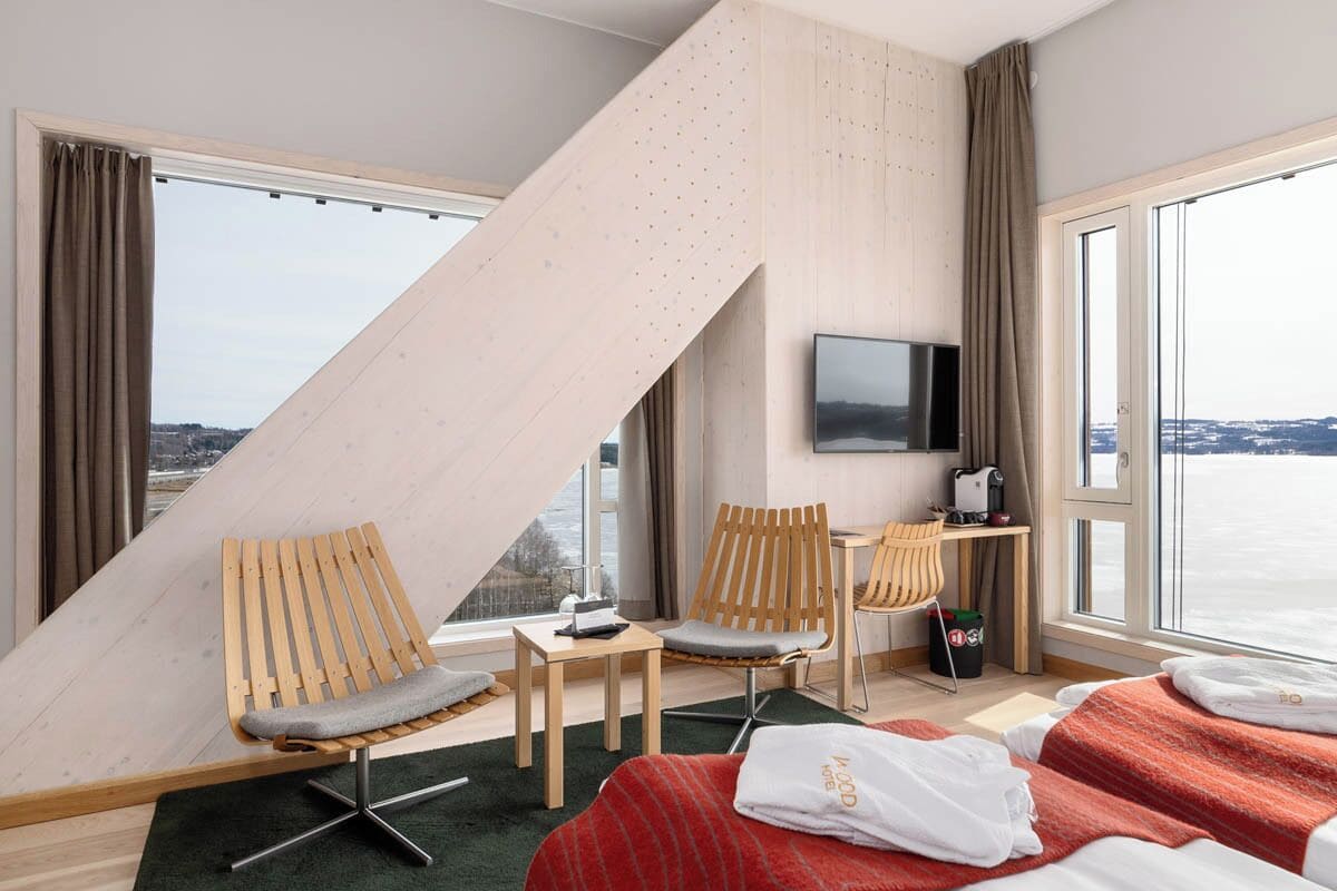 Quarto de hotel com teto inclinado, duas cadeiras de madeira, uma pequena mesa, uma cama com roupa de cama vermelha, uma televisão e vista para uma paisagem nevada.