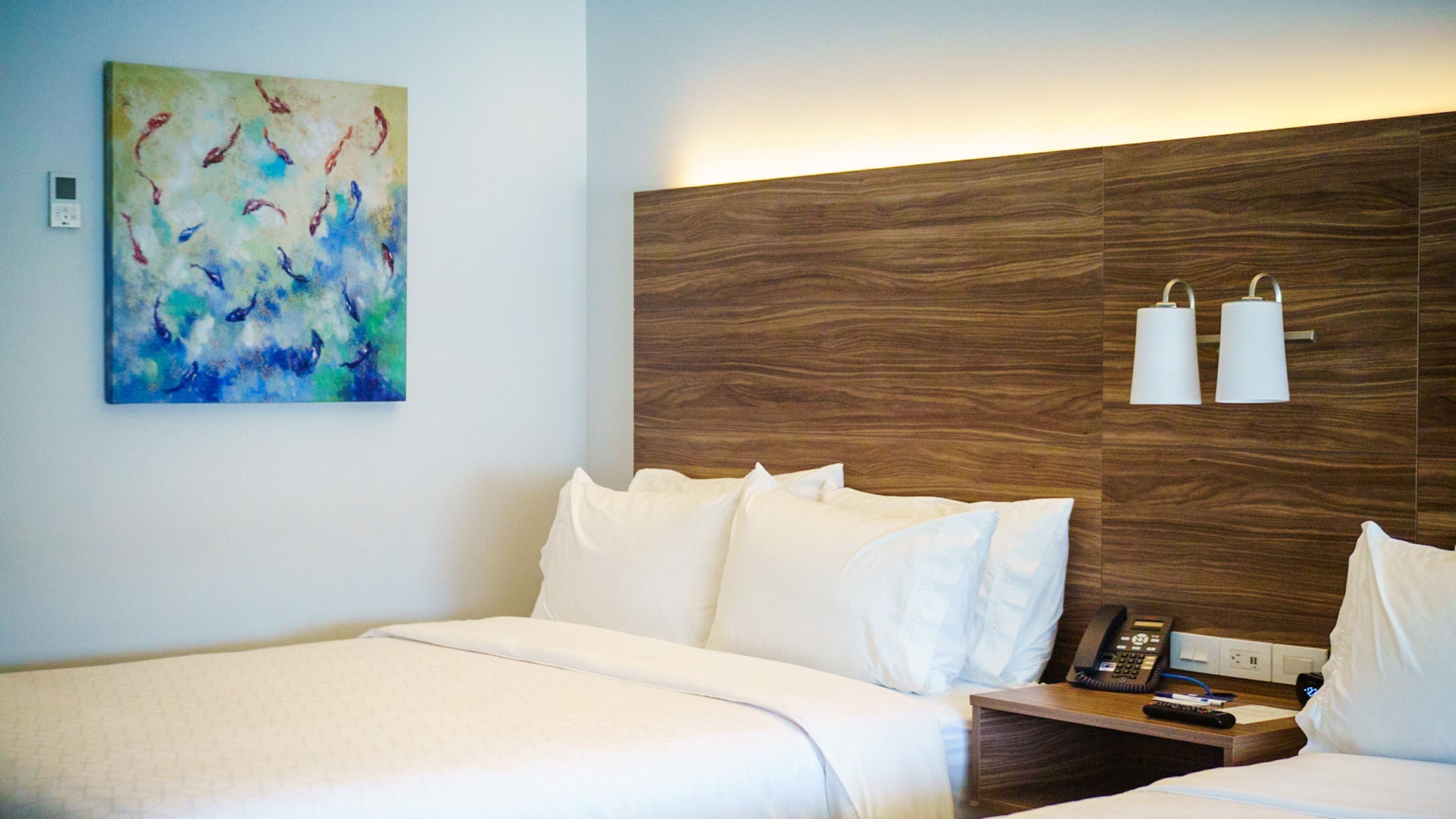 Quarto de hotel com cama, mesa de cabeceira, telefone e luminária fixada na parede.