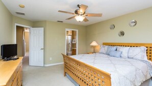 Quarto com cama de madeira, ventoinha no teto, televisão sobre um aparador e porta dando acesso a outro cômodo.