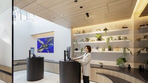 Uma área de recepção moderna com uma recepcionista em uma mesa, cercada por prateleiras com plantas e uma grande pintura abstrata na parede.
