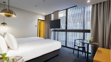 Quarto de hotel com cama, escrivaninha, cadeira, televisão e janela com cortinas.