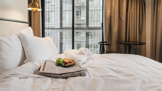 Quarto de hotel com cama, janela com cortinas, uma mesinha e luminária suspensa.