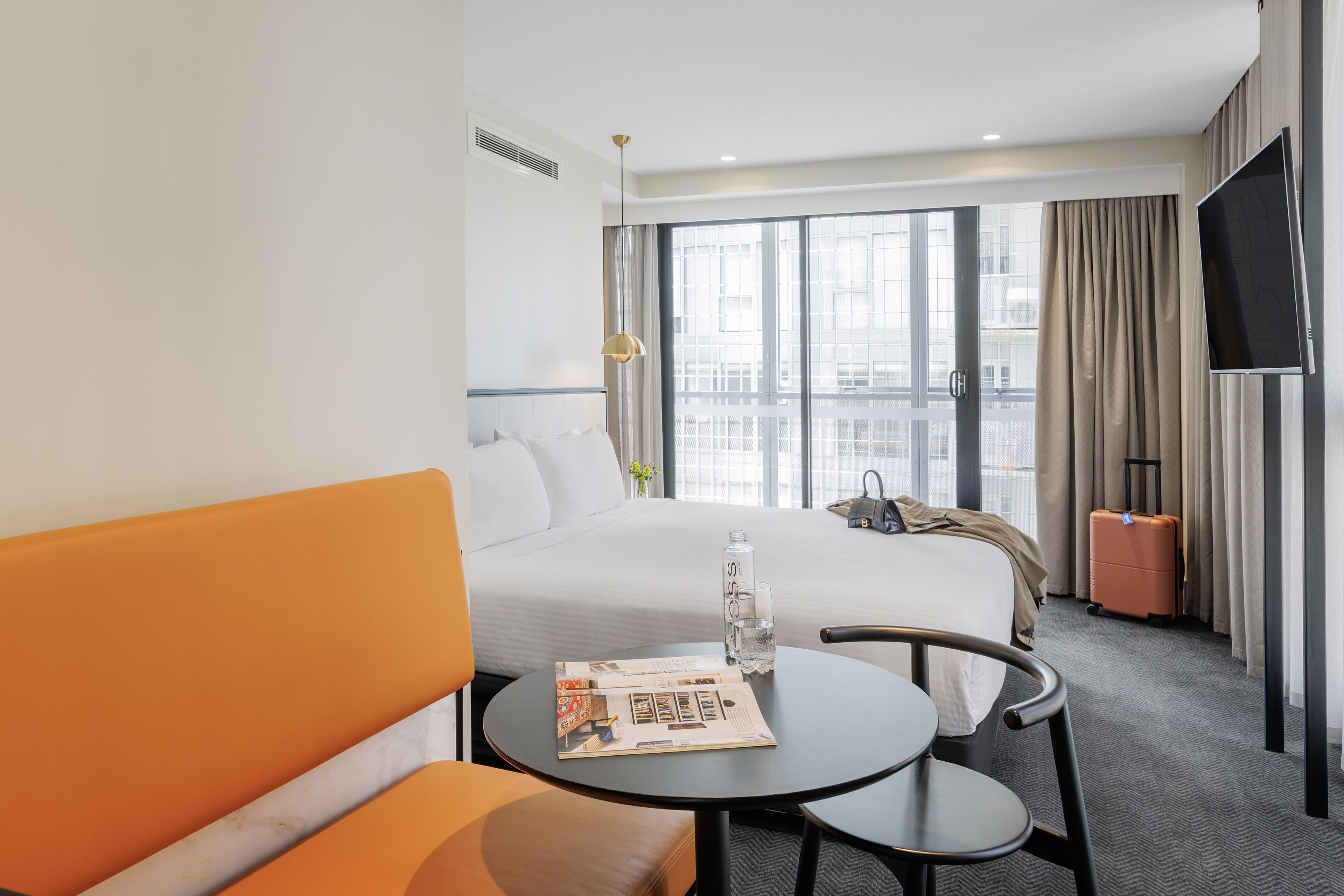 Quarto de hotel com cama, mesa pequena, cadeira, televisão e janela com cortinas.
