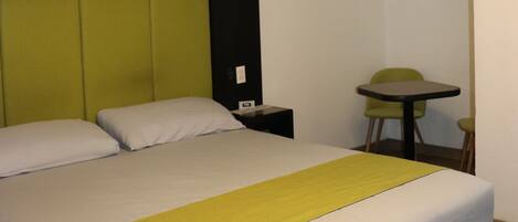 Quarto de hotel com uma cama, mesa de cabeceira, uma cadeira verde e uma pequena mesa.