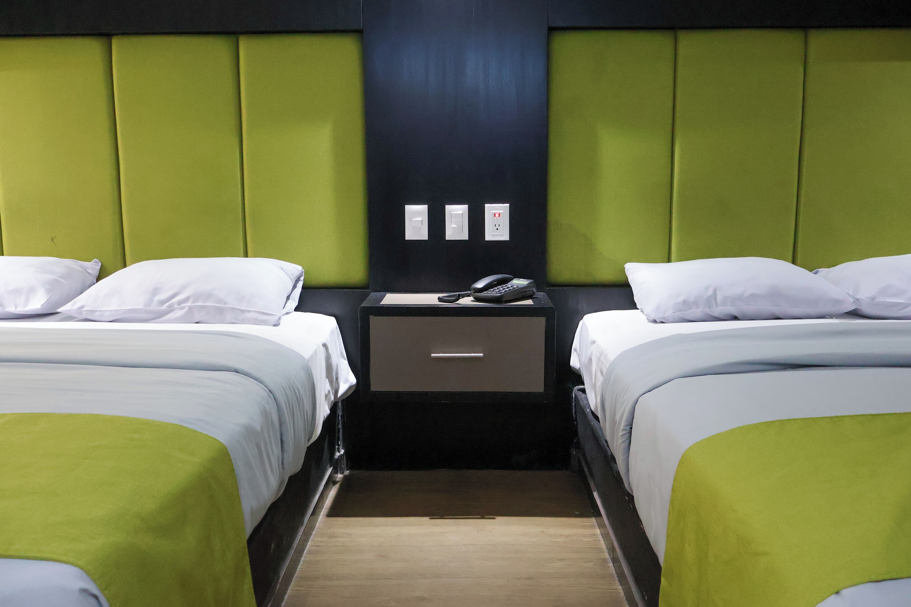 Quarto de hotel com duas camas, cada uma com roupa de cama branca e cabeceiras verdes. Há uma mesa de cabeceira com um telefone e uma gaveta.