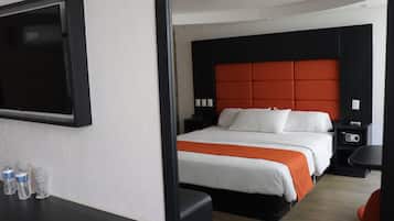 Quarto de hotel com uma cama grande, uma TV fixada na parede, uma mesa de cabeceira com abajur e uma pequena mesa com água engarrafada.