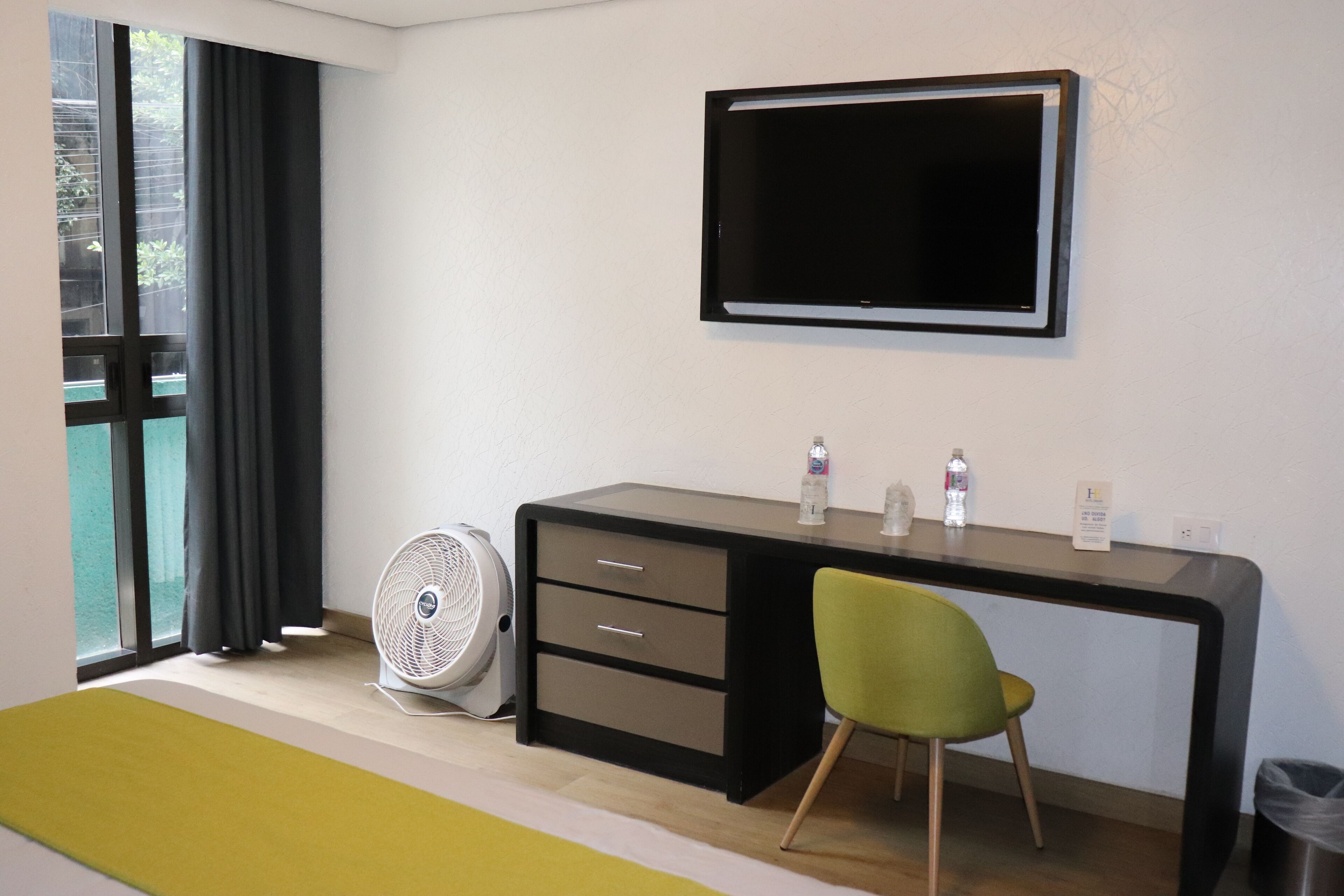 Quarto com escrivaninha, cadeira, televisão, ventoinha e janela com cortinas.