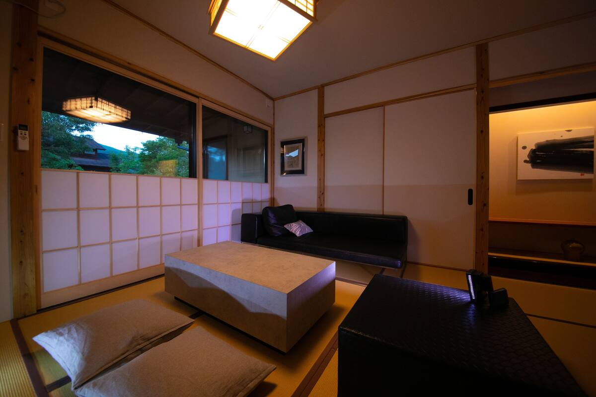 Um quarto japonês tradicional com tatames, uma mesa baixa, um sofá e uma porta de correr com vista para o verde lá fora.