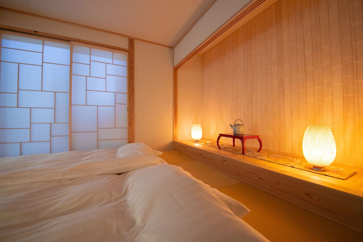 Quarto de hotel com cama, mesa de cabeceira, luminária e porta deslizante com padrão geométrico.