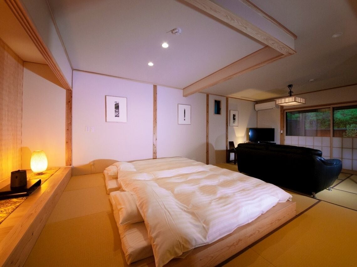 Quarto com cama, escrivaninha com luminária, sofá preto e televisão.