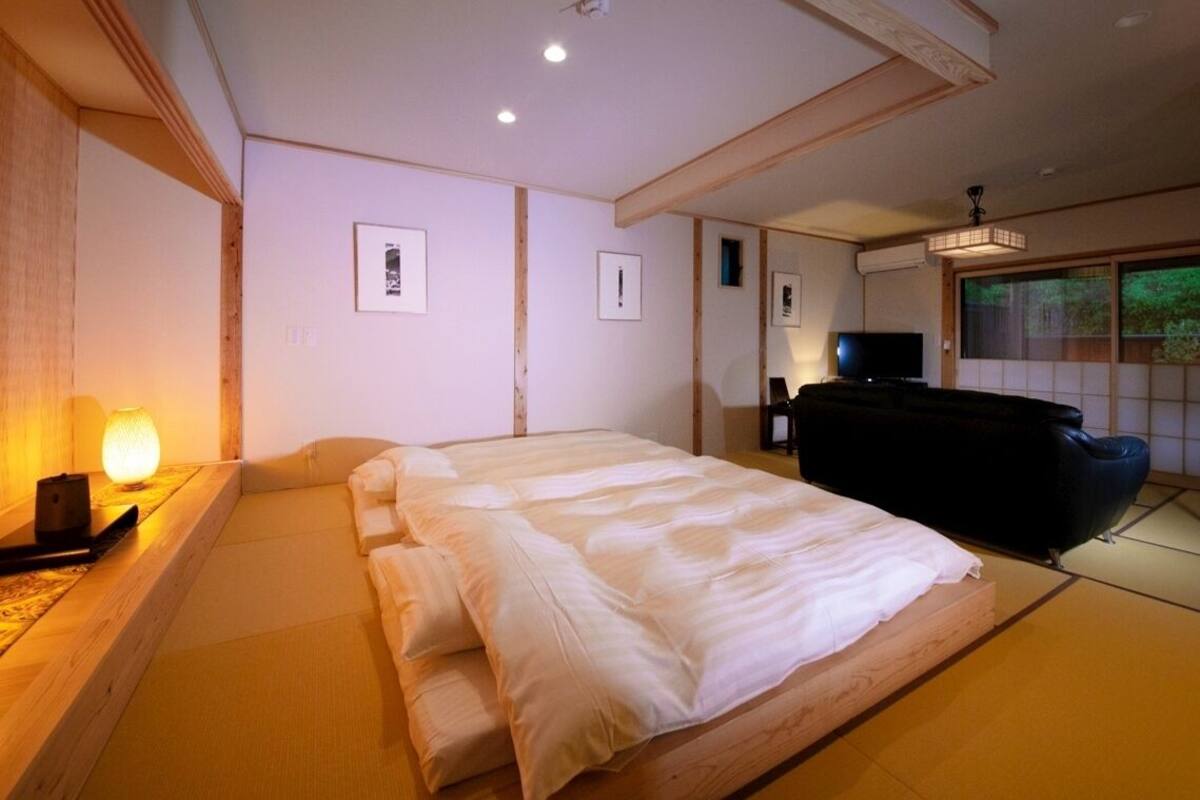 Quarto com cama, escrivaninha com luminária, sofá preto e televisão.