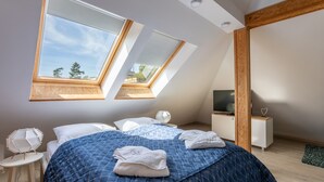 Quarto com claraboia, cama com colcha azul, travesseiros brancos, mesa de cabeceira e televisão.
